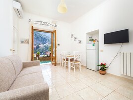 Uma sala de estar com sofá, mesa de jantar e cadeiras, televisão na parede e porta que dá para uma varanda com vista para uma cidade e montanhas.