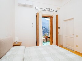 Quarto com cama, mesa de cabeceira, janela com cortinas e vista para montanhas.