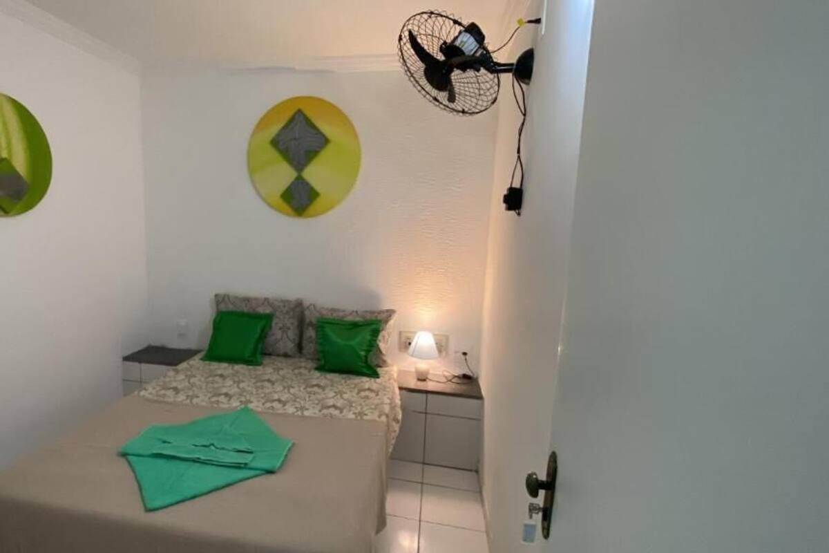 Quarto com cama, almofadas verdes, mesa de cabeceira com abajur e duas ventoinhas fixadas na parede.