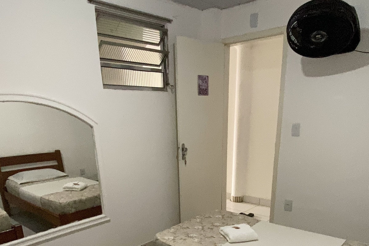 Quarto pequeno e limpo, com duas camas de solteiro, um espelho, uma janela e uma porta.