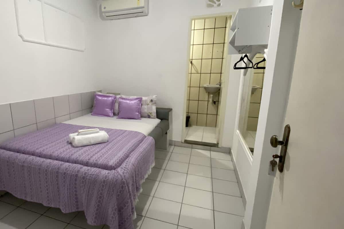 Quarto pequeno e limpo, com uma cama, chuveiro e porta branca.