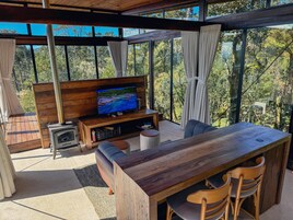 Uma área moderna de estar ao ar livre com uma mesa e cadeiras de madeira, uma televisão sobre um suporte de madeira, e vista para uma paisagem arborizada através de grandes janelas.