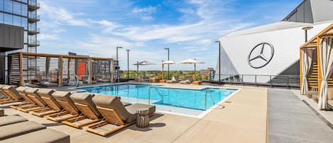 Uma área de piscina no terraço com espreguiçadeiras e guarda-sóis, o logotipo da Mercedes e um edifício moderno ao fundo.