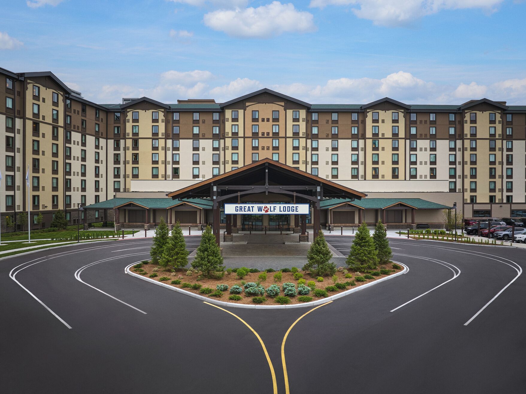 Um hotel com vários andares, entrada central, estacionamento e área ajardinada com árvores.