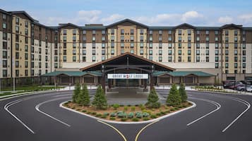 Um hotel com vários andares, entrada central, estacionamento e área ajardinada com árvores.