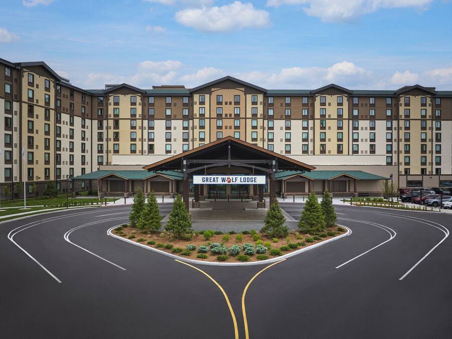 Um hotel com vários andares, entrada central, estacionamento e área ajardinada com árvores.
