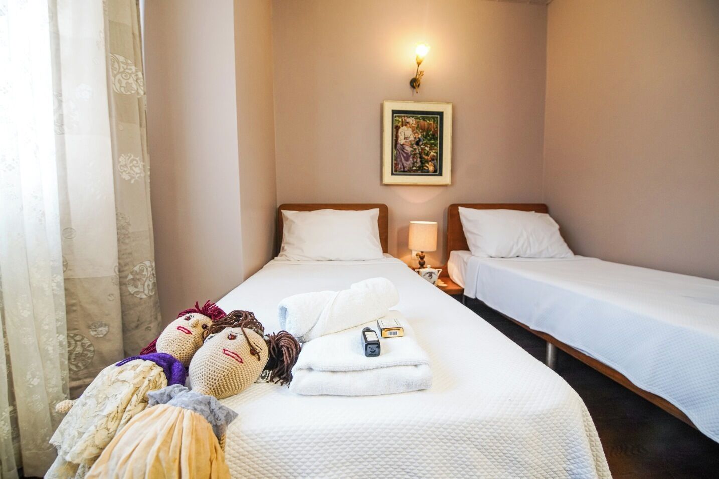 Quarto de hotel com duas camas, uma mesa de cabeceira com abajur, um quadro na parede e uma janela com cortinas.