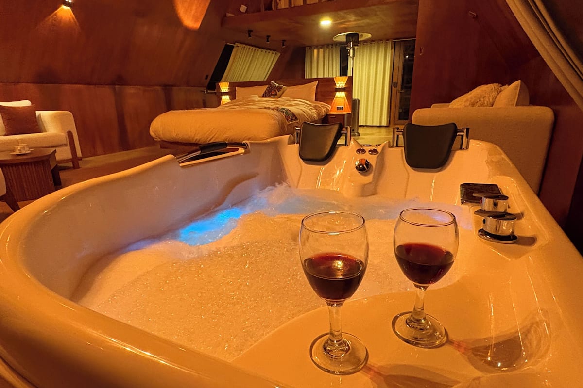 Uma banheira de hidromassagem com duas taças de vinho tinto, decoração em madeira e vista para o quarto.