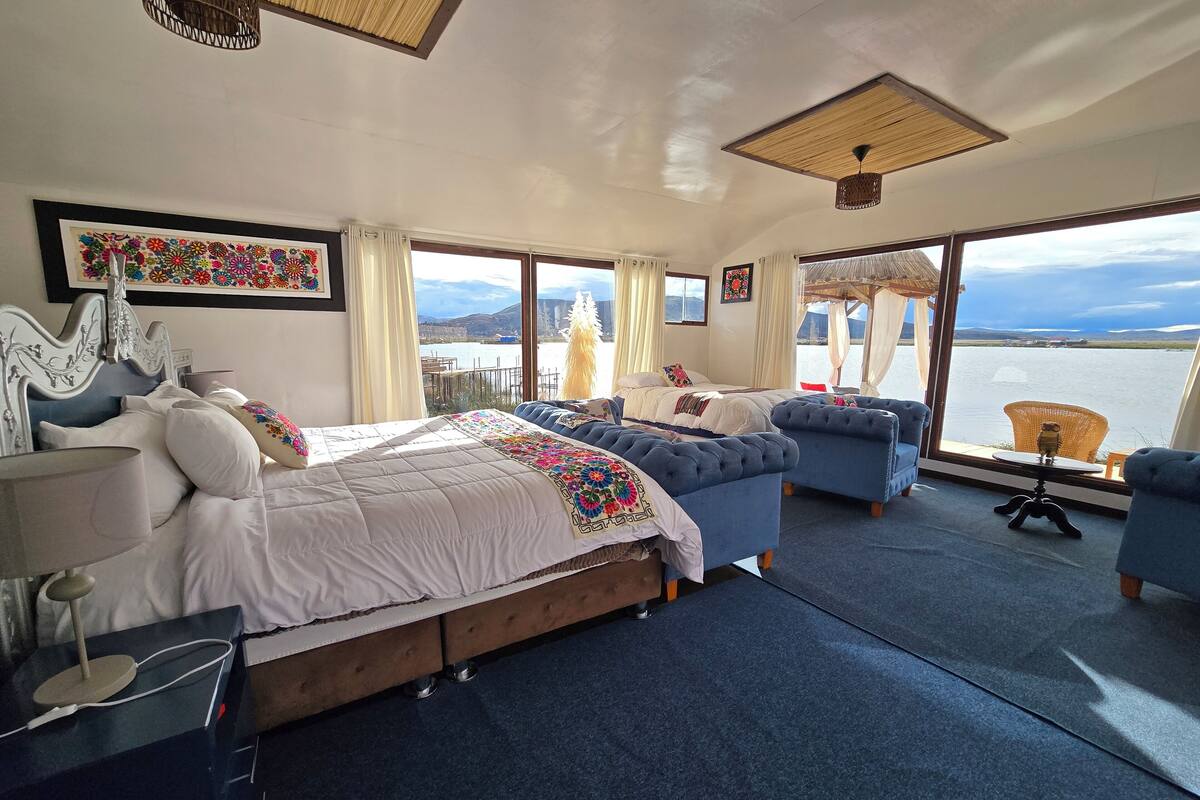Quarto com uma cama grande, dois sofás azuis, uma mesinha e vista para um lago.