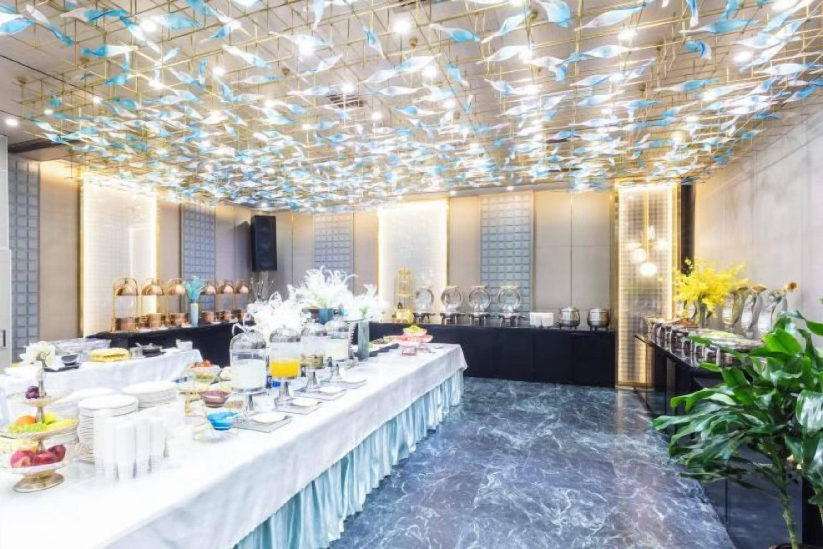 Uma sala de banquetes com uma longa mesa posta para um evento formal, piso de mármore e luminárias pendentes no teto.