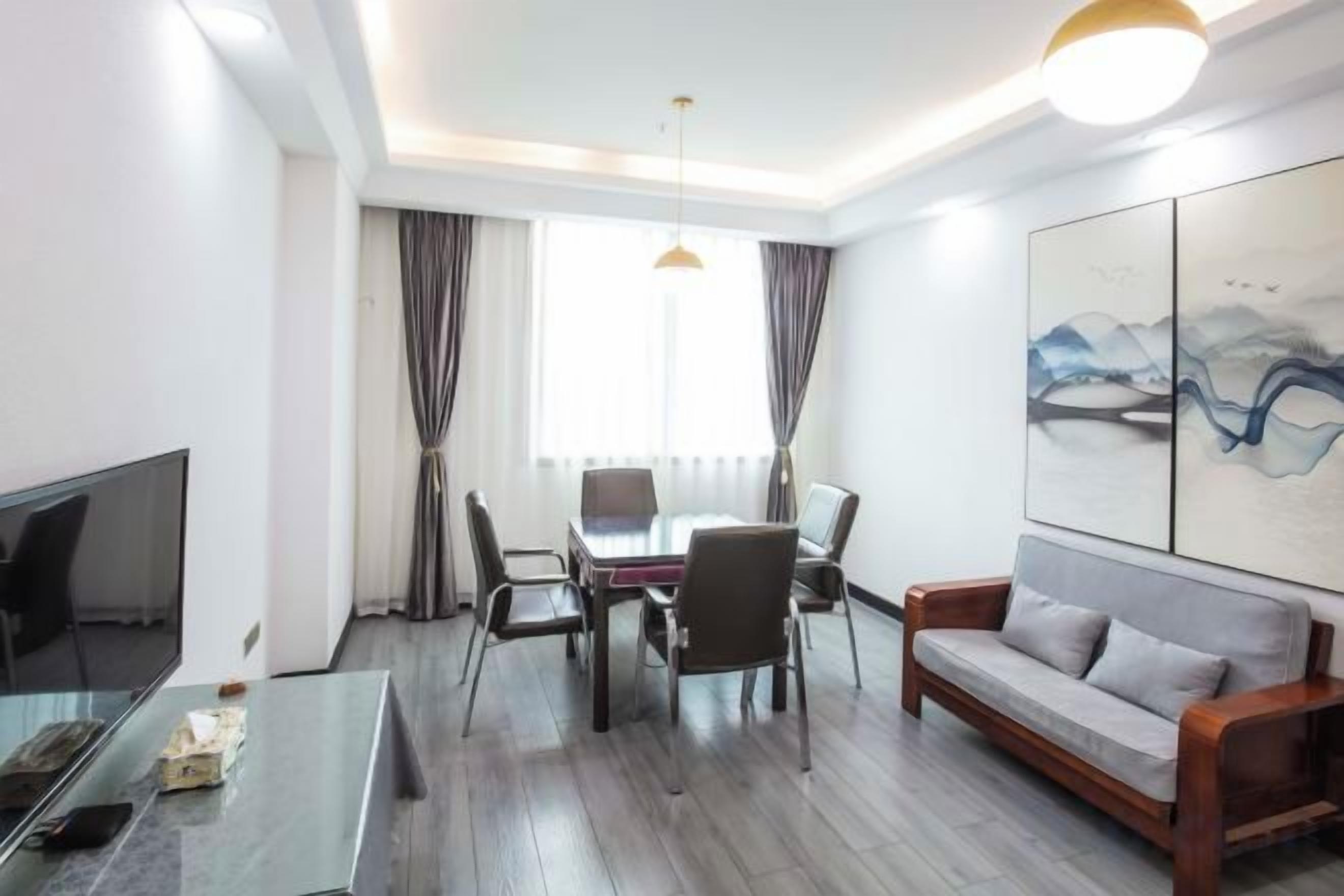Quarto de hotel com televisão, mesa de jantar com cadeiras, sofá e uma janela grande com cortinas.