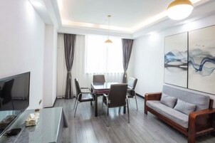 Quarto de hotel com televisão, mesa de jantar com cadeiras, sofá e uma janela grande com cortinas.