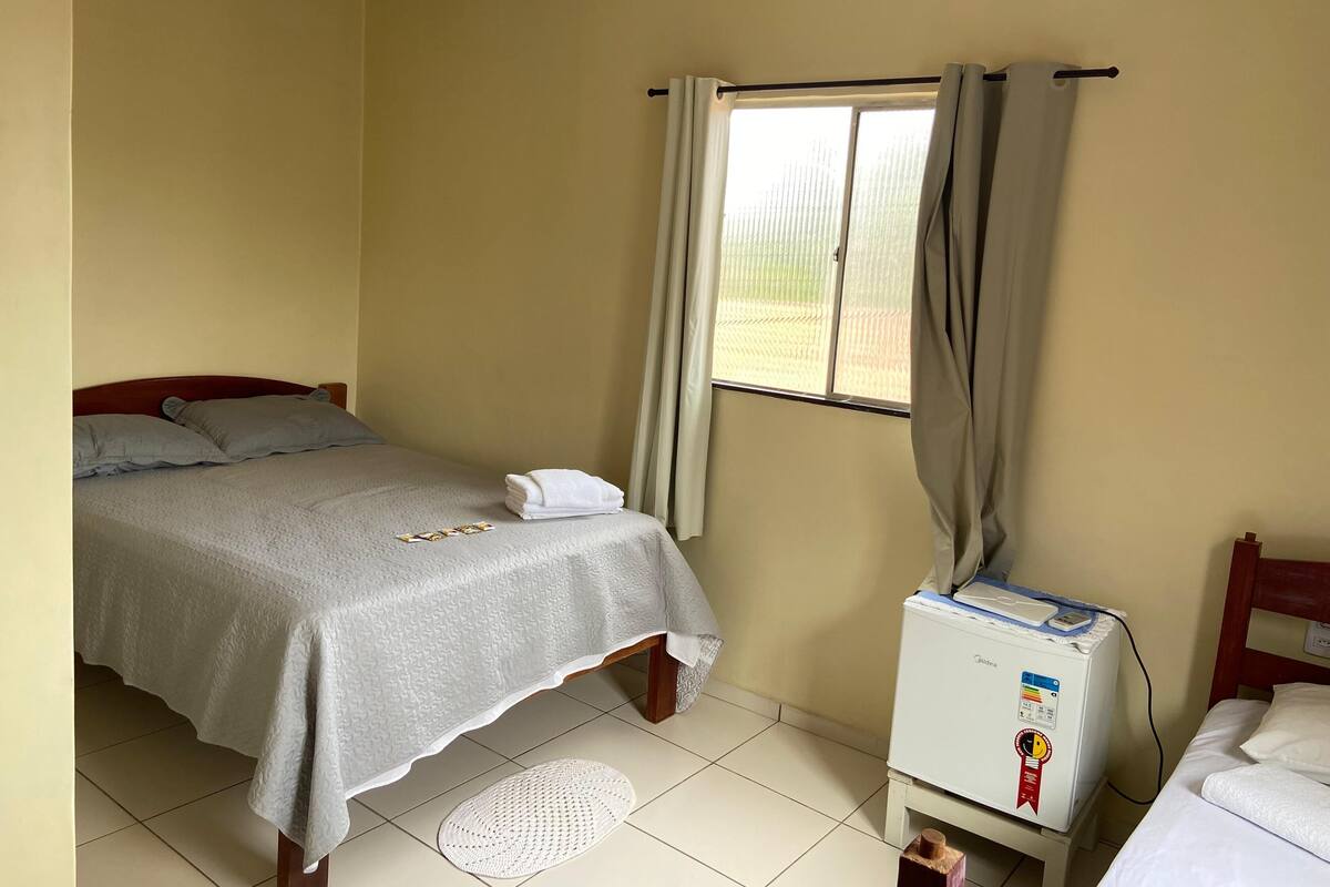 Quarto pequeno e limpo, com uma cama, uma mesa pequena com um frigorífico, janela com cortinas e chão em mosaico.