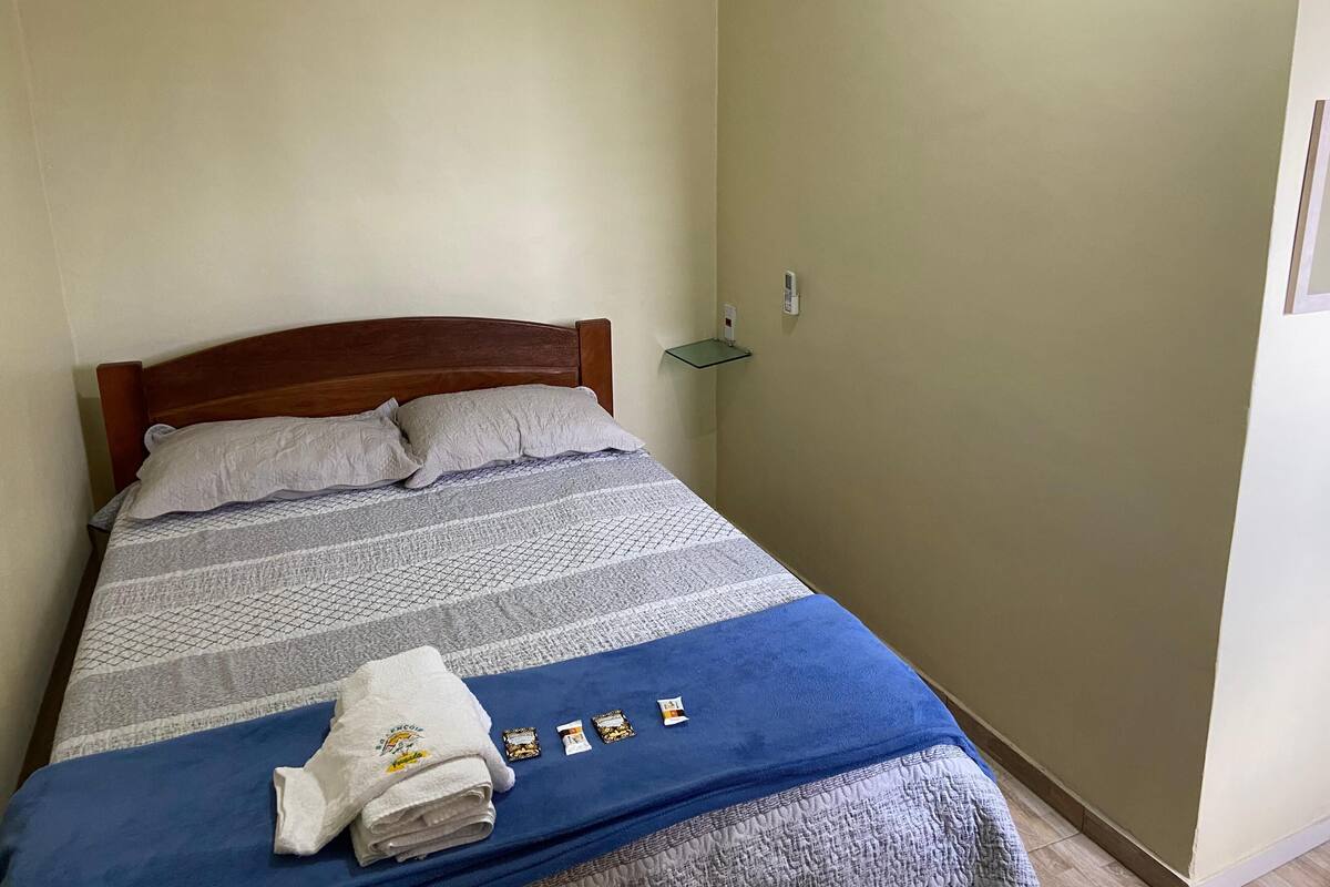 Cama bem arrumada com manta azul e toalha dobrada, mesa de cabeceira com prateleira de vidro e interruptor de luz na parede.