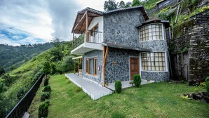 Uma casa de dois andares com fachada de pedra, uma varanda e vista para as colinas ao redor.