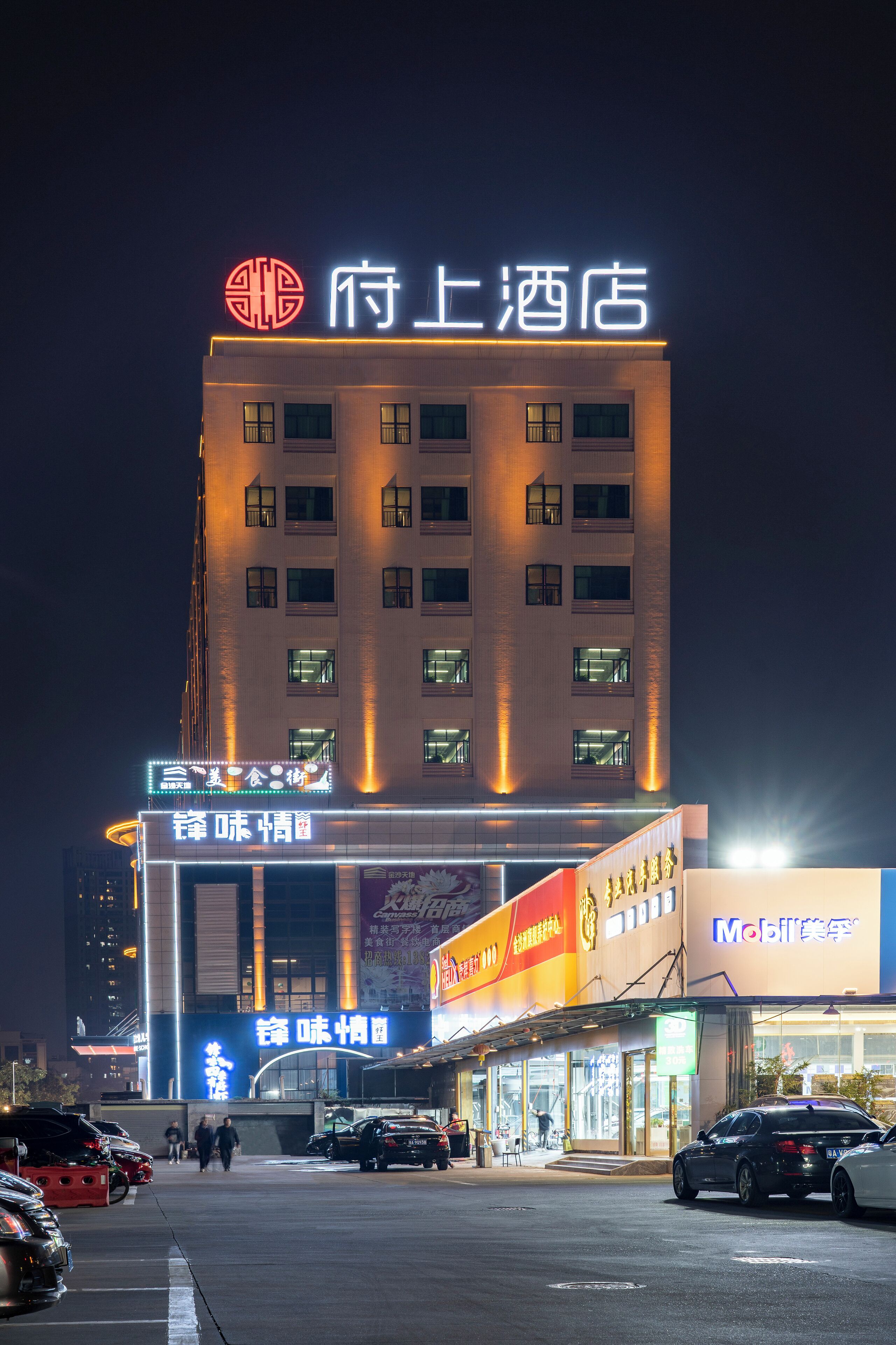 Um hotel com uma placa grande no topo, várias lojas no nível da rua e carros estacionados em primeiro plano.
