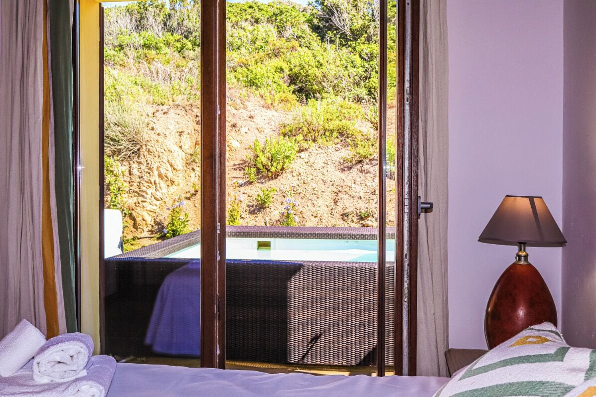 Quarto de hotel com cama, janela e vista para piscina e vegetação, além de uma luminária.