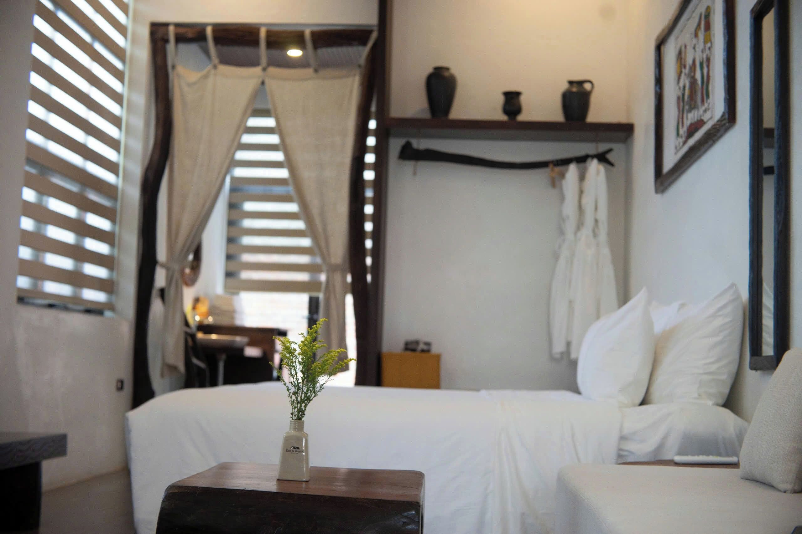 Um quarto com cama, mesa de cabeceira com um vaso, janela com persianas, prateleira com objetos decorativos e um quadro na parede.