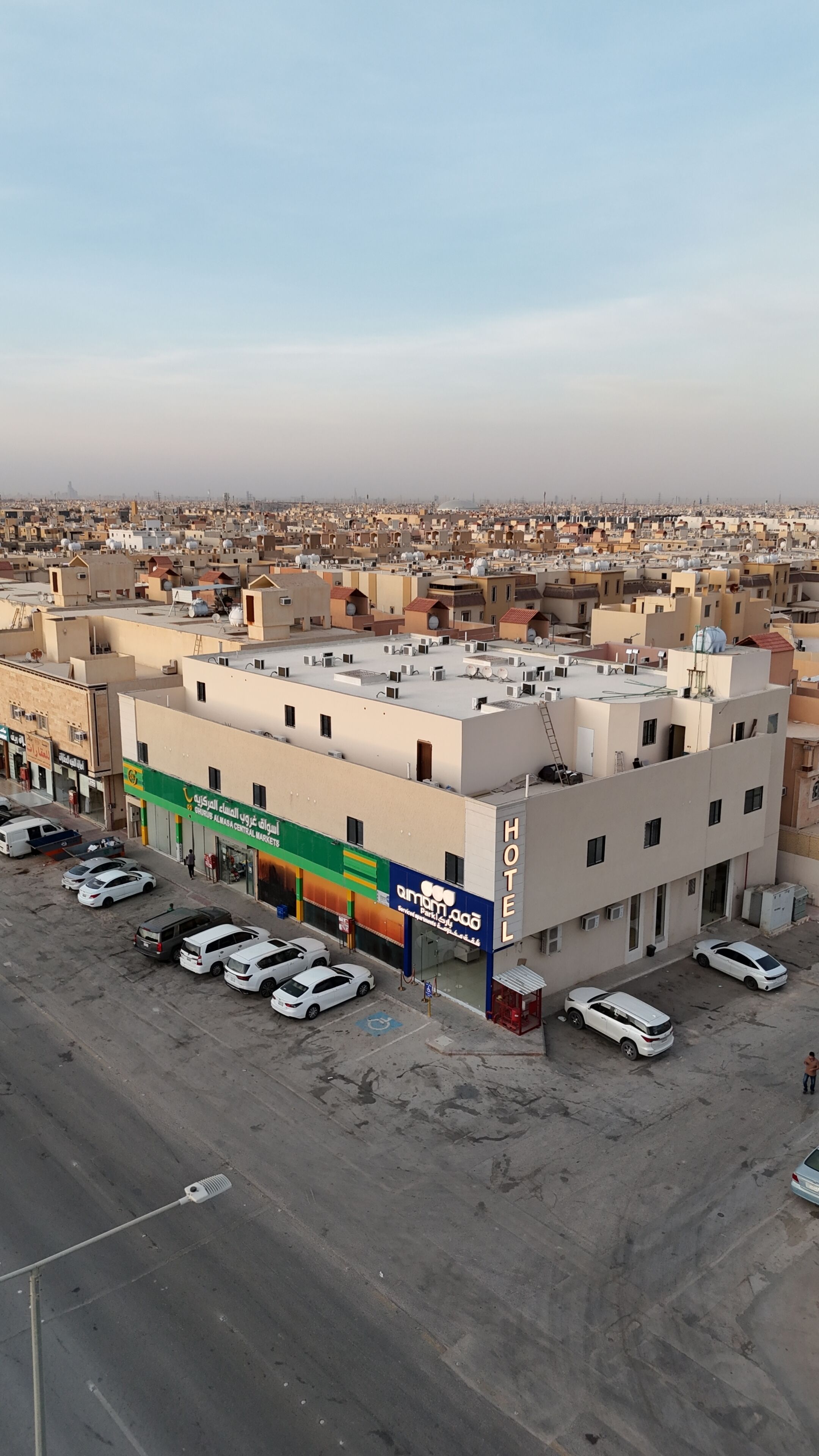 Um hotel com telhado plano, vários espaços de estacionamento e uma placa a indicar 'Alma Central Markets'.