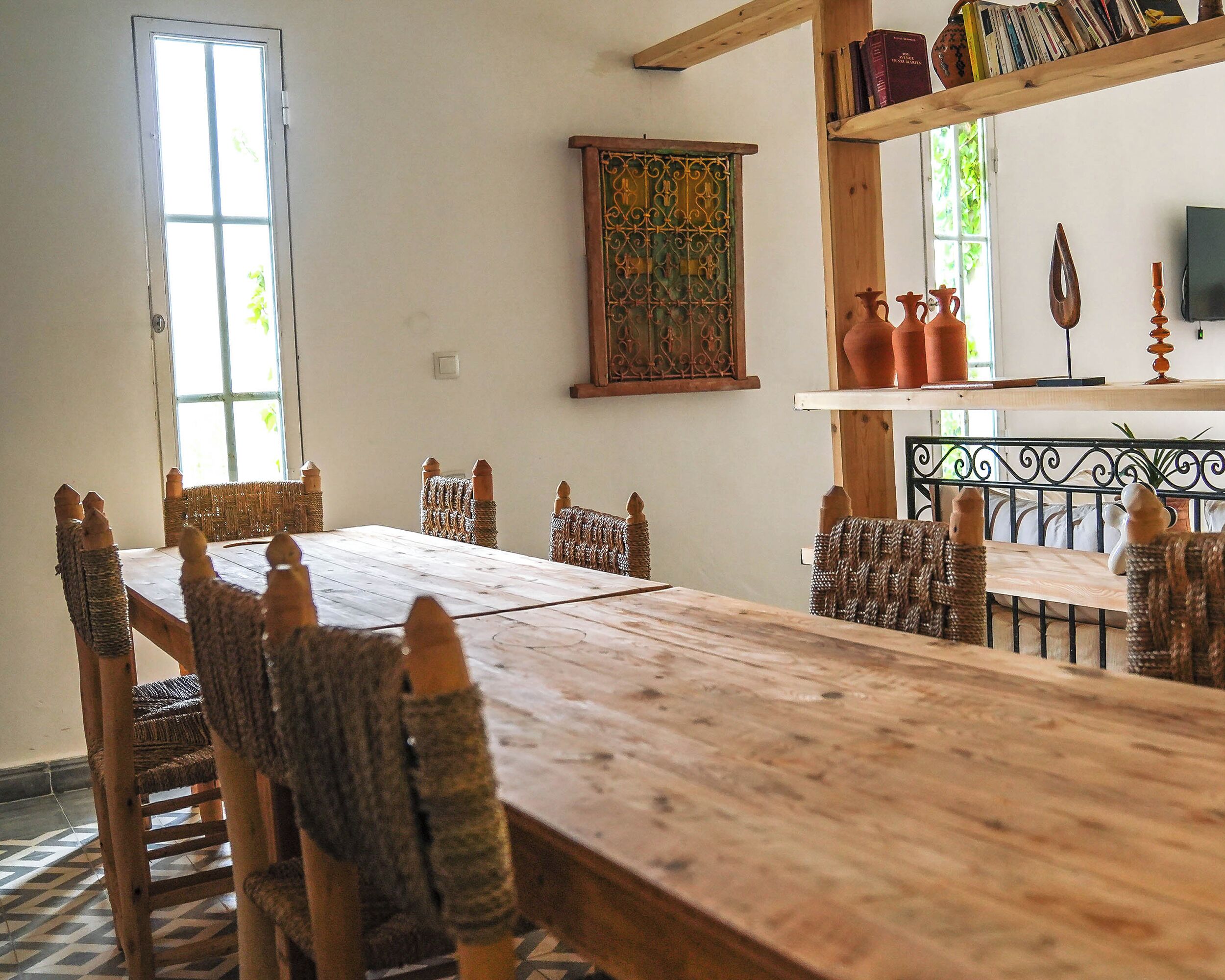 Uma área de refeições com mesa e cadeiras de madeira, uma estante com livros e objetos decorativos, e uma janela com luz natural.