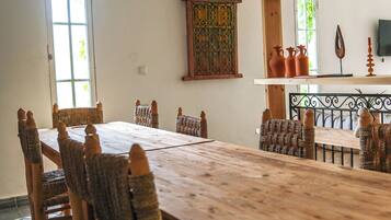 Uma área de refeições com mesa e cadeiras de madeira, uma estante com livros e objetos decorativos, e uma janela com luz natural.