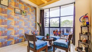 Quarto com parede quadriculada, uma janela grande com cortinas, uma pequena mesa redonda com uma planta e uma cadeira de madeira com almofada.