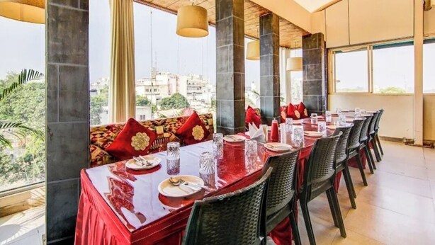 Uma sala de jantar com uma mesa comprida posta para vários convidados, janelas amplas com vista para edifícios e vegetação, e teto alto com vigas aparentes.