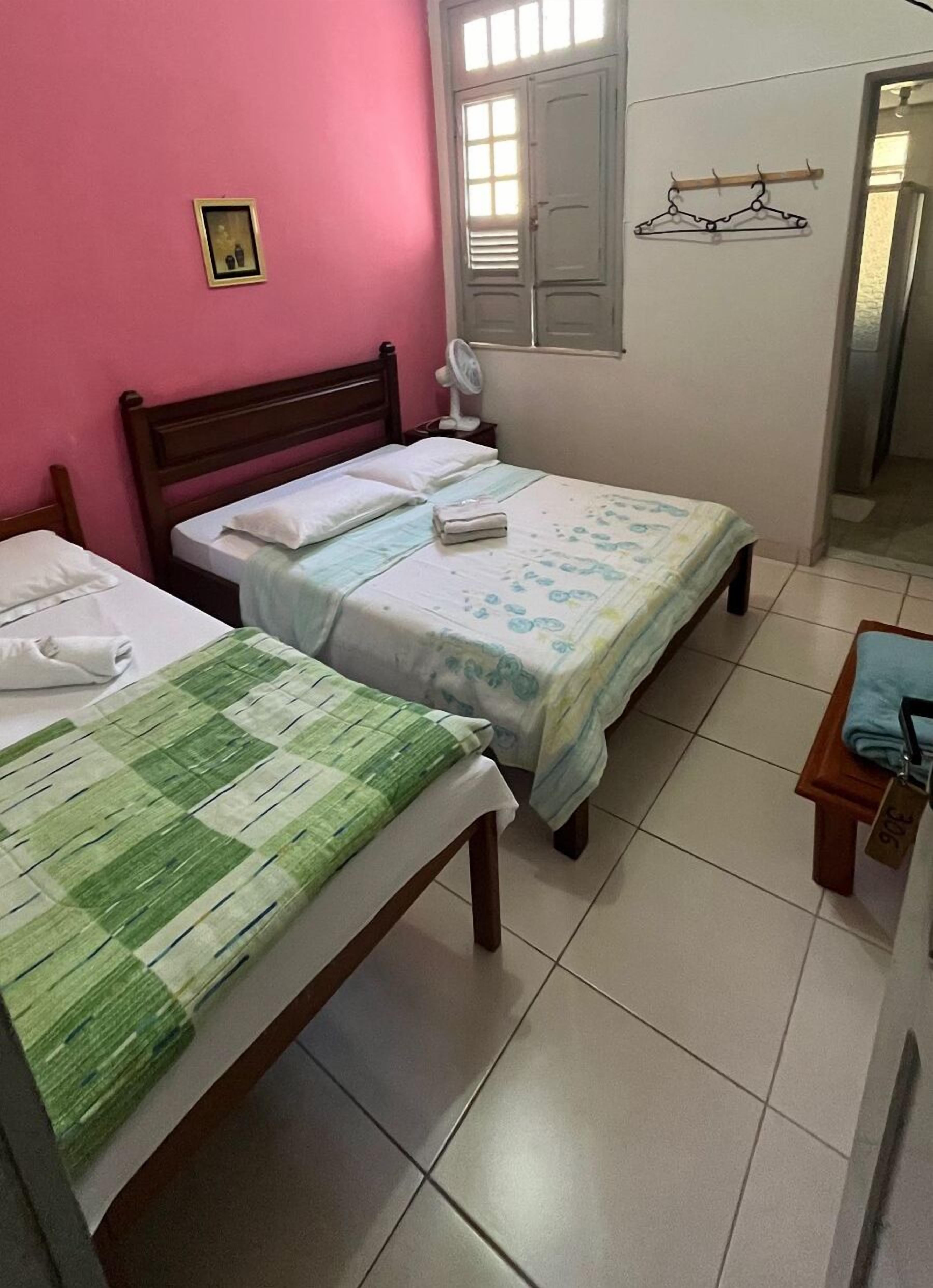 Quarto com duas camas, cabeceira de madeira, mesa de cabeceira com ventilador, janela com persianas e cabideiro na parede.