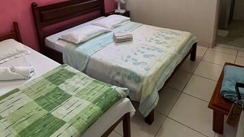 Quarto com duas camas, cabeceira de madeira, mesa de cabeceira com ventilador, janela com persianas e cabideiro na parede.