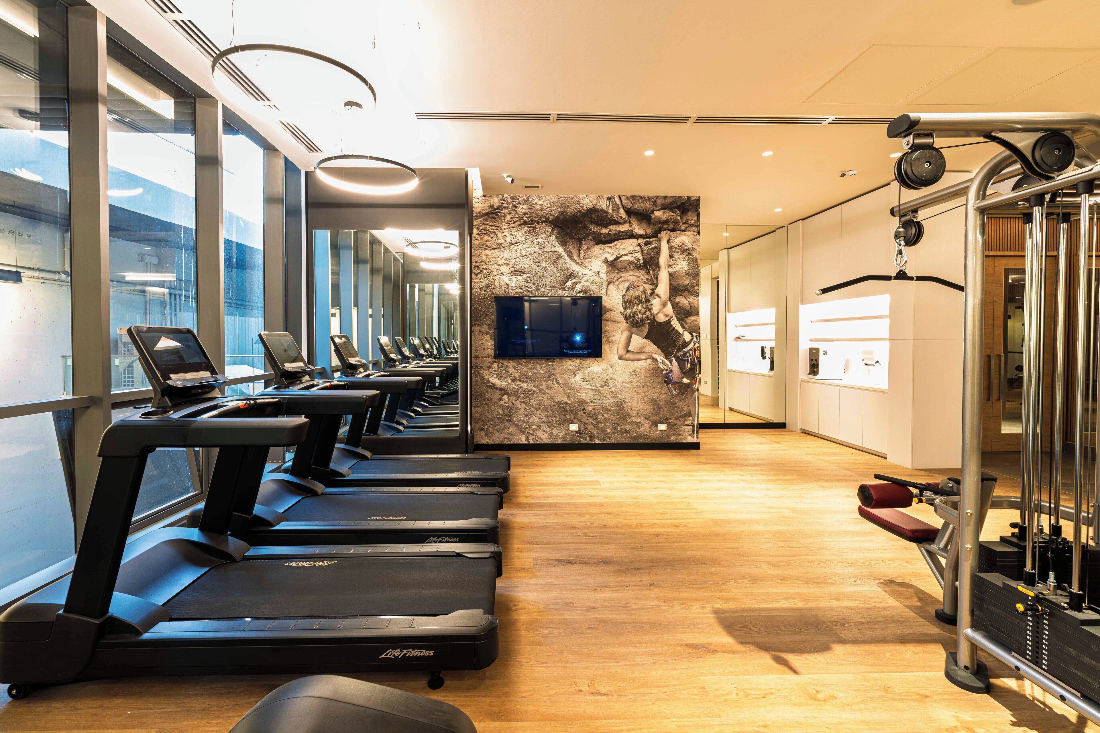 Uma academia com esteiras, uma televisão fixada na parede e equipamentos de exercícios.