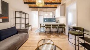 Uma sala de estar moderna com um sofá, uma área de jantar com banquetas e uma cozinha ao fundo.