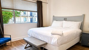 Quarto moderno com uma cama grande, um banquinho, uma mesa de cabeceira e vista para edifícios pela janela.