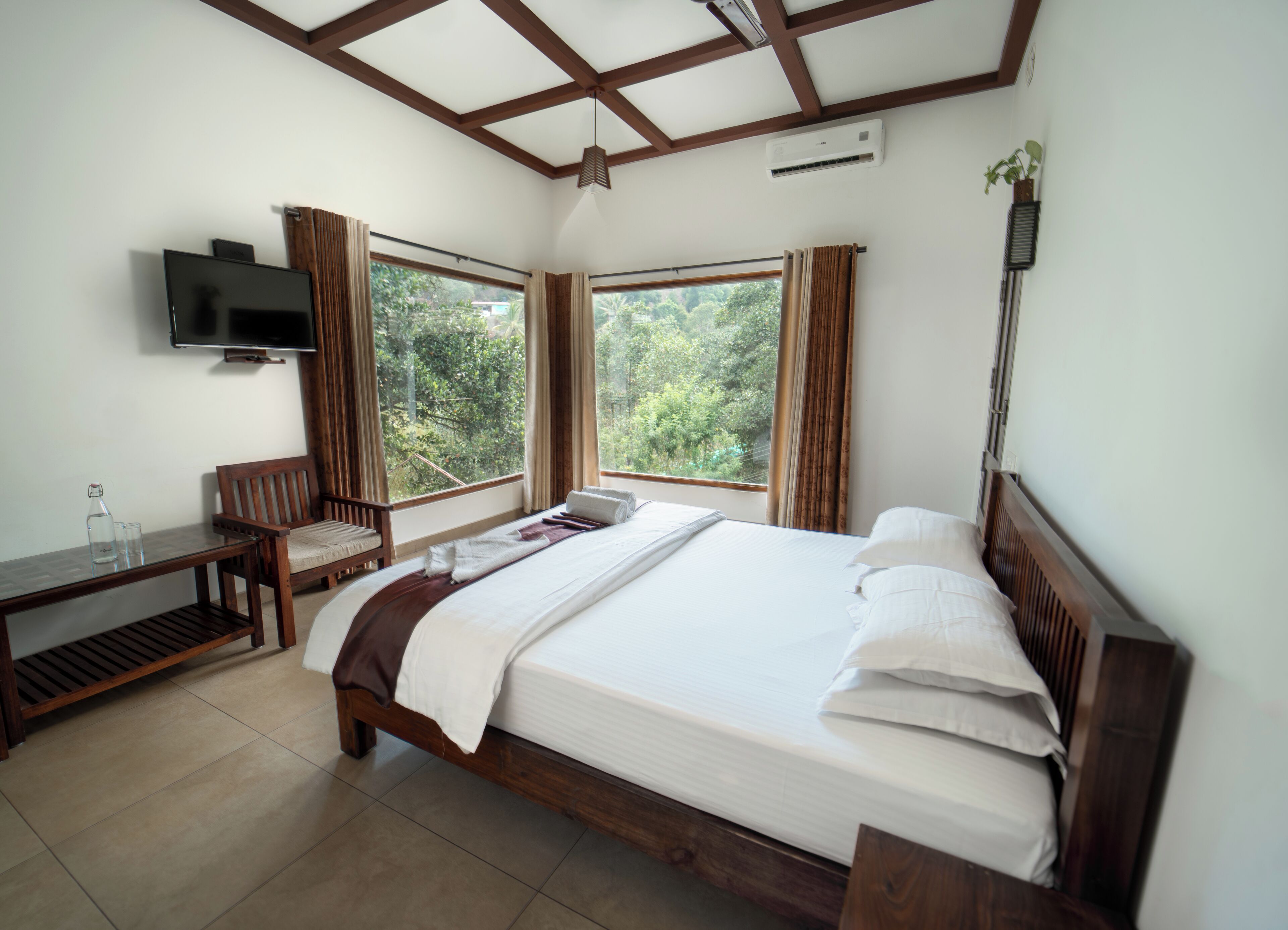 Quarto com cama, escrivaninha, televisão e janelas amplas com cortinas.