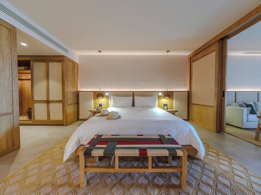 Quarto espaçoso com uma cama grande, abajures, um banquinho de madeira e vista para uma varanda.