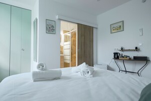 Quarto de hotel com cama, guarda-roupa de madeira, área de chuveiro e uma pequena mesa com chaleira e chávenas.