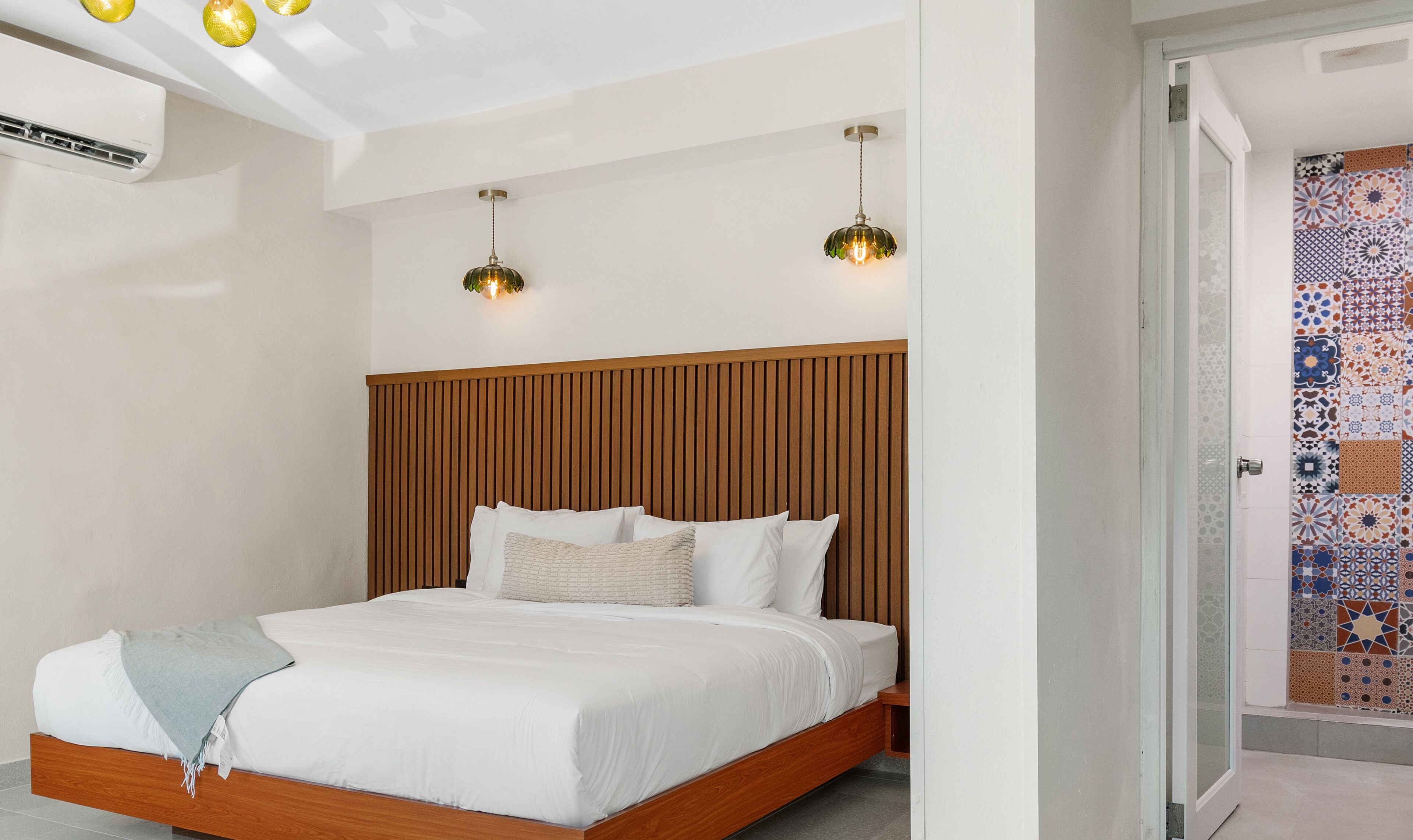 Quarto com cabeceira de madeira, cama com roupa de cama branca, duas luzes pendentes e parede com azulejos estampados.
