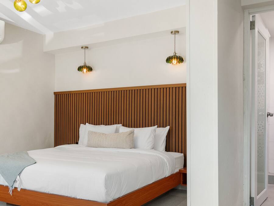 Quarto com cabeceira de madeira, cama com roupa de cama branca, duas luzes pendentes e parede com azulejos estampados.