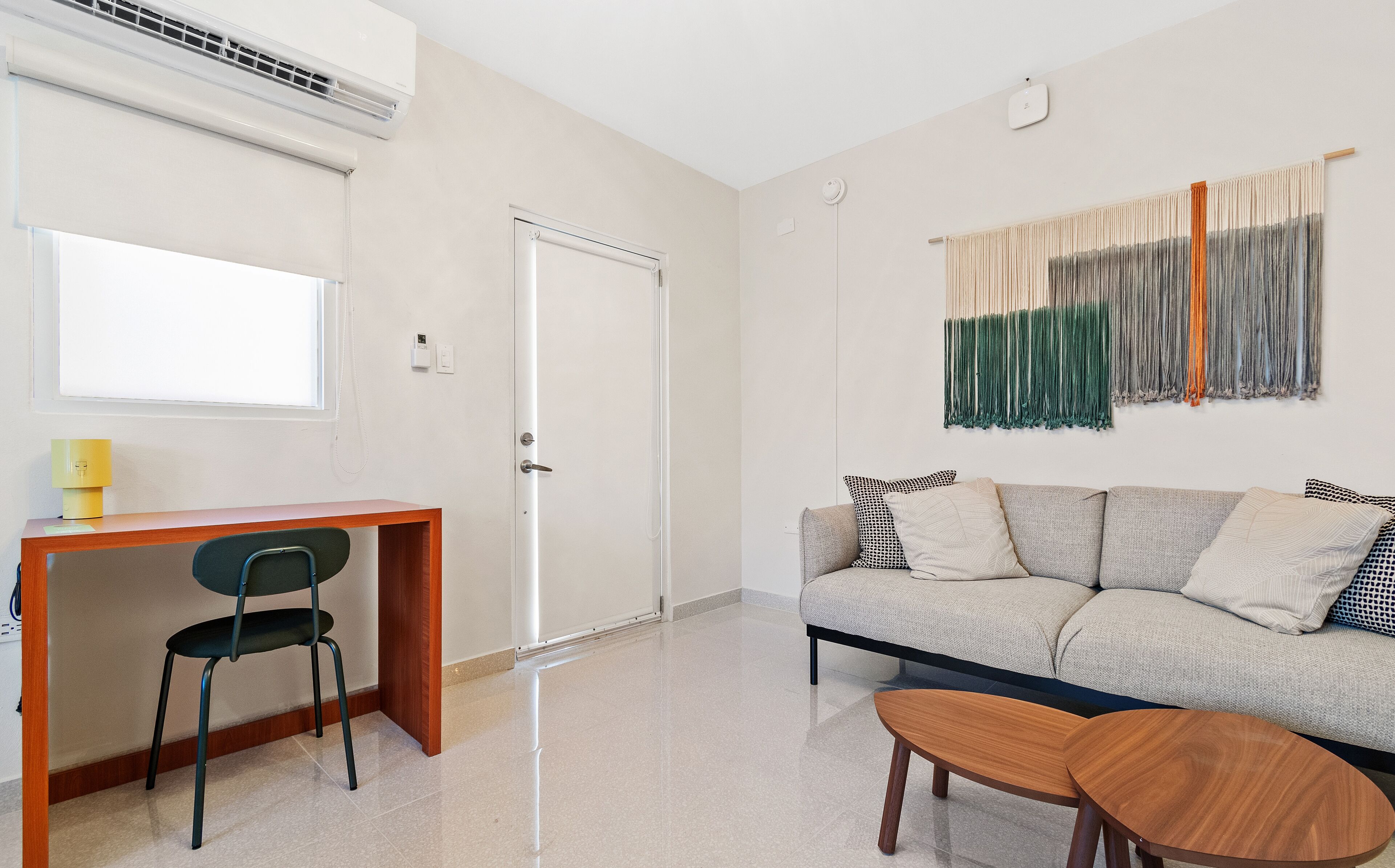 Quarto moderno com escrivaninha de madeira, uma cadeira verde, um sofá com almofadas estampadas e uma tapeçaria na parede.