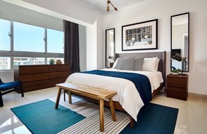 Quarto moderno com uma cama grande, um banco, cômoda e vista da cidade por grandes janelas.
