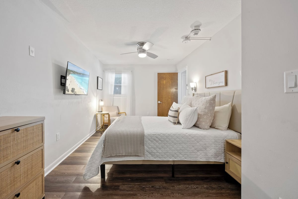 Quarto com cama, mesas de cabeceira, ventilador de teto, televisão e janela.