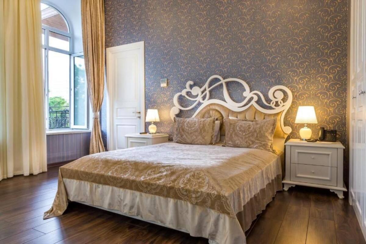 Quarto com cama grande, cabeceira decorada, duas mesas de cabeceira com luminárias, uma janela com cortinas e papel de parede estampado.