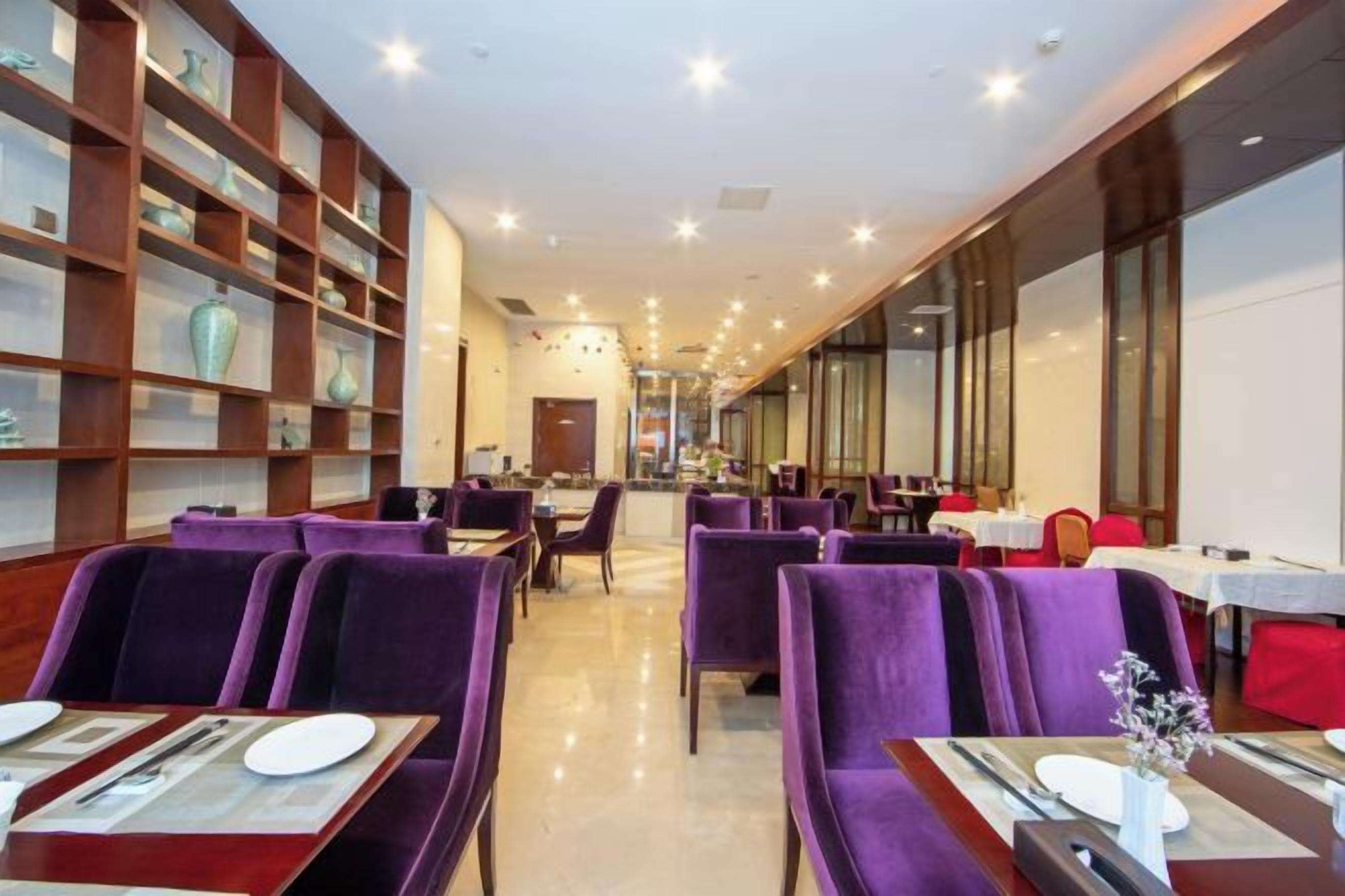 Uma sala de jantar de hotel com cadeiras estofadas em roxo, uma passagem central e uma mesa comprida com um vaso de flores.