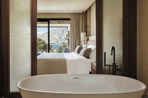 Quarto de hotel moderno com uma banheira grande, cama com roupa de cama branca e vista para uma paisagem montanhosa através de uma janela ampla.