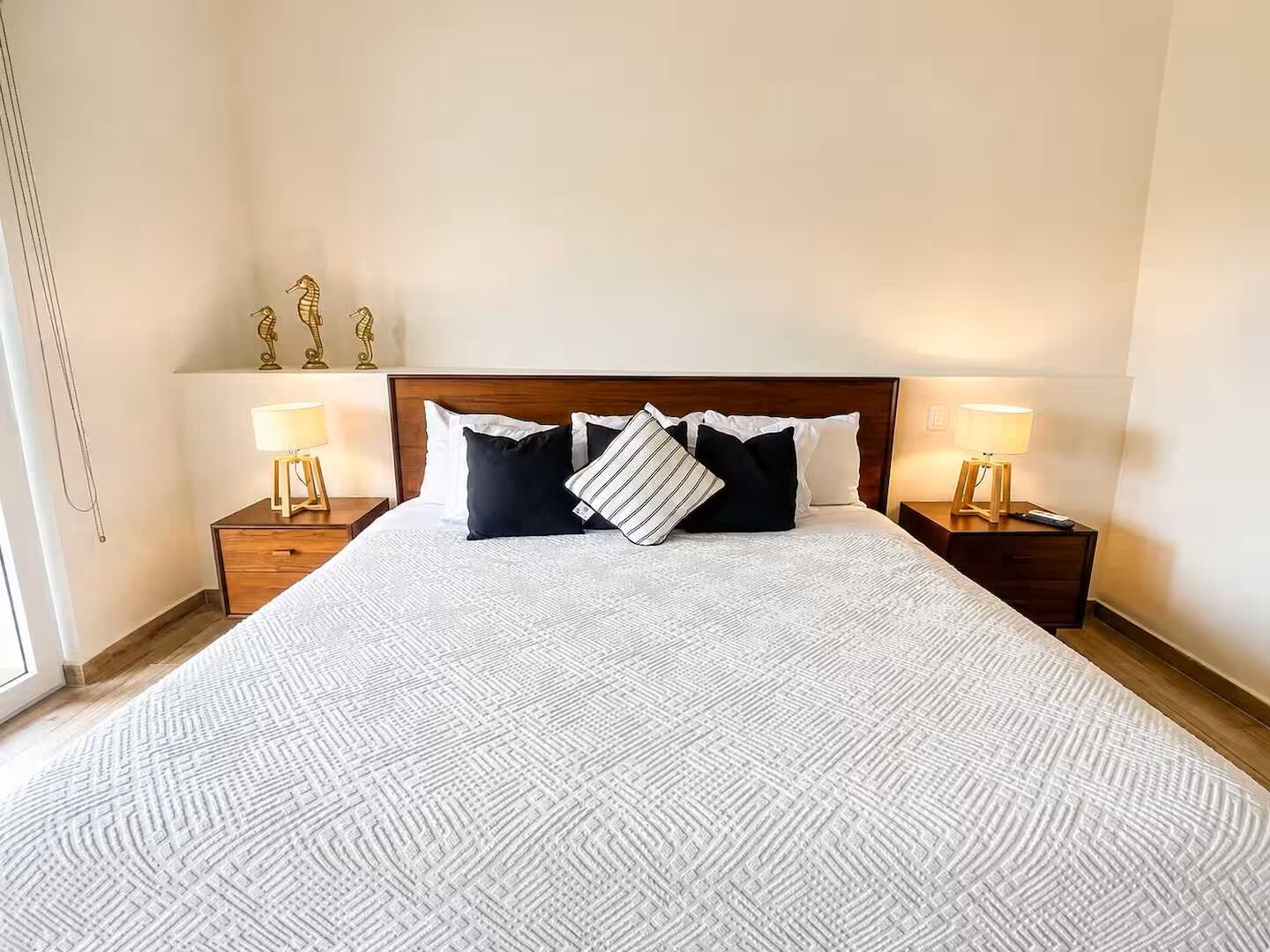 Uma cama bem arrumada com um cobre-leito estampado, com duas mesas de cabeceira com abajures, num quarto com paredes lisas e chão de madeira.