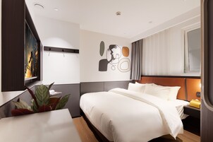 Quarto de hotel moderno com uma cama grande, televisão fixada na parede e uma planta no canto.