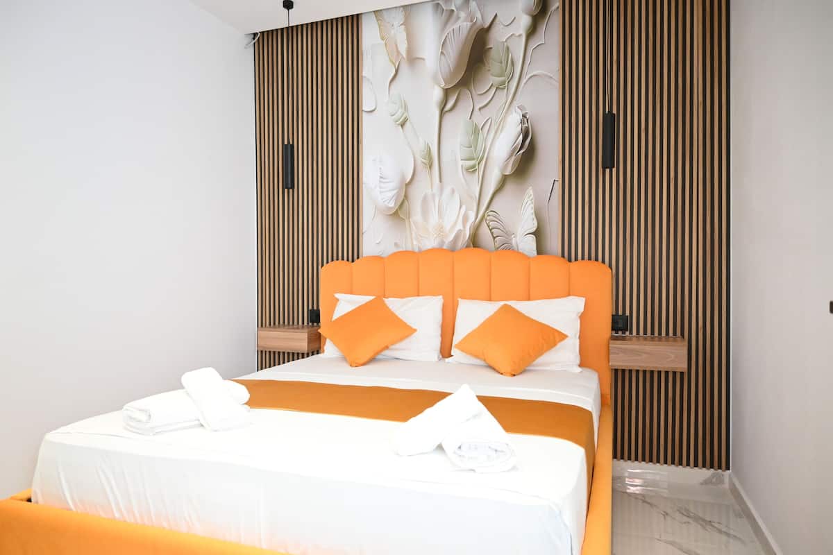 Quarto de hotel moderno com cama grande, cabeceira laranja, roupa de cama branca e um painel decorativo na parede.