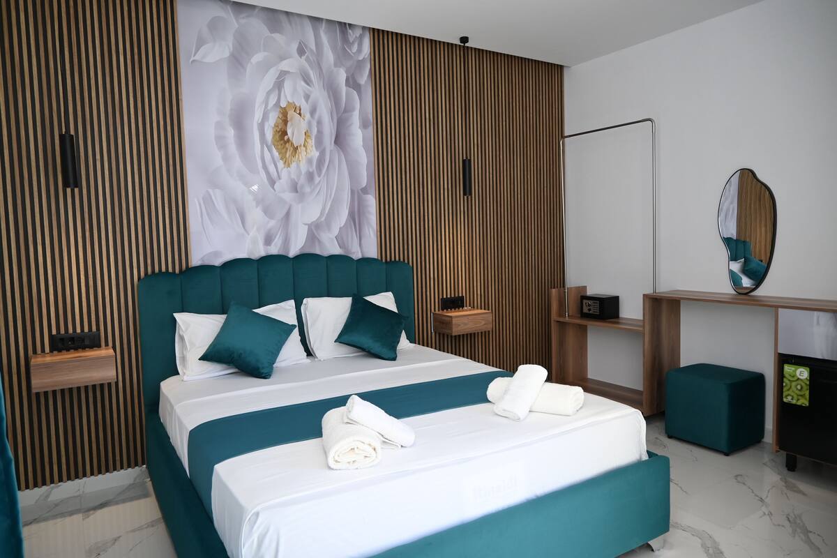Quarto de hotel moderno com uma cama grande, roupa de cama verde-água, painel de parede de madeira, uma grande obra de arte floral, uma mesinha de cabeceira com rádio e um espelho na parede.