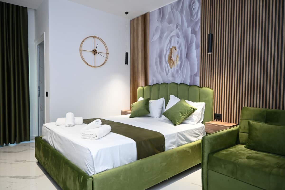 Quarto de hotel moderno com uma cama grande, mobiliário estofado em verde, um relógio de parede e um mural floral.