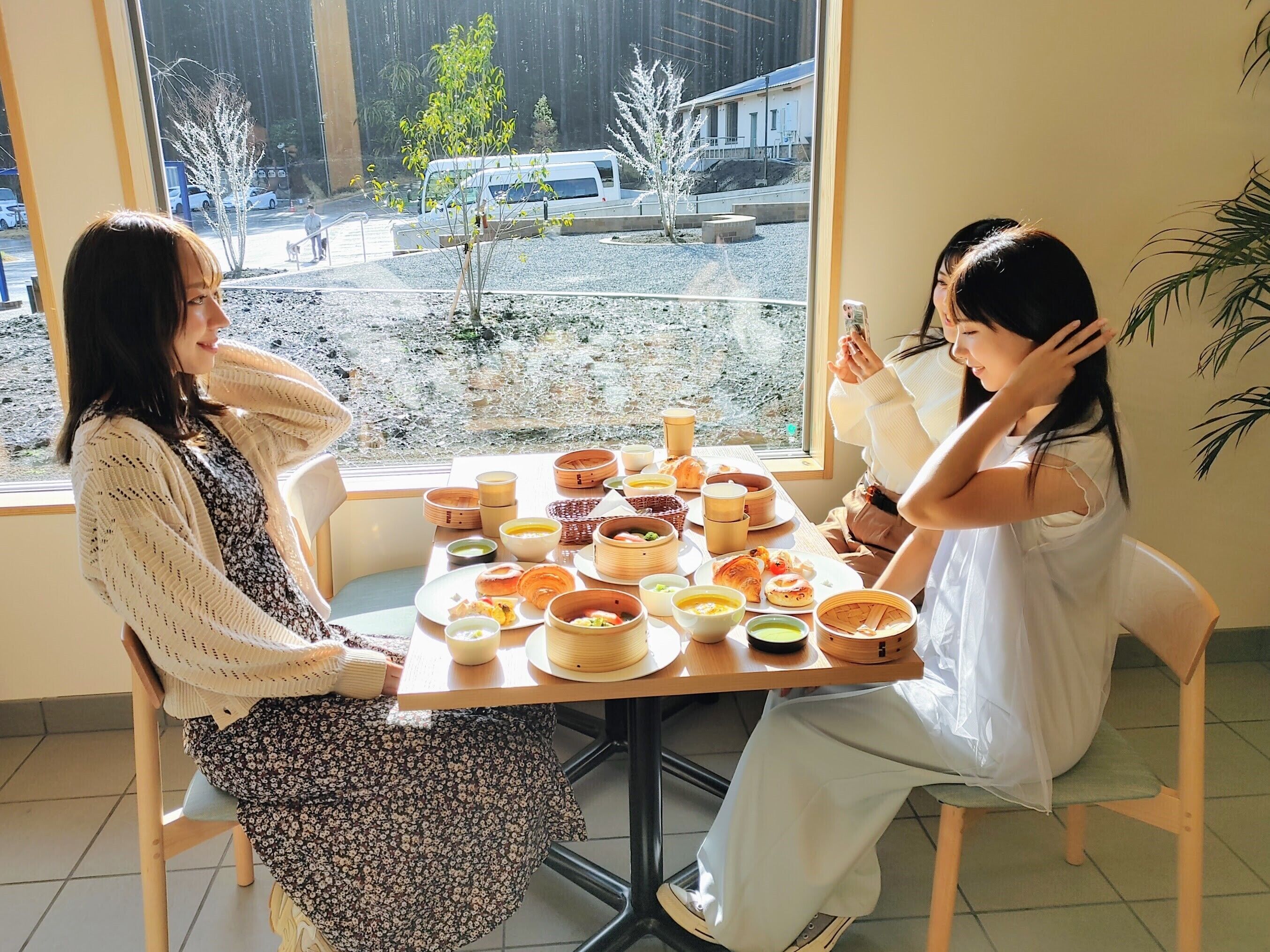 Duas pessoas sentadas à mesa com diversos pratos, vista pela janela de um estacionamento e edifícios, e um vaso de planta.