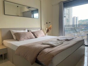 Quarto de hotel moderno com uma cama grande, uma mesa de cabeceira, um espelho e vista para a cidade através de amplas janelas.